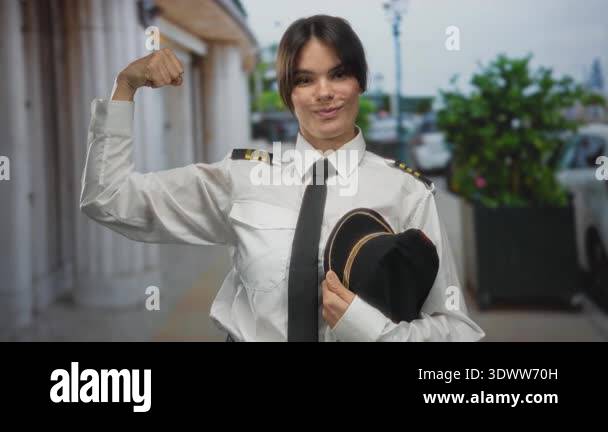 Young hispanic woman pilot in uniform flexing arm confidently on city ...