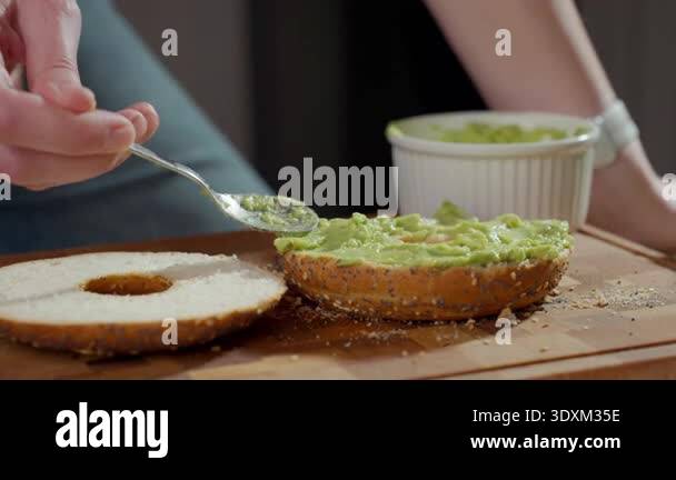 Hands spreading mashed avocado on bagel with spoon on wooden cutting ...