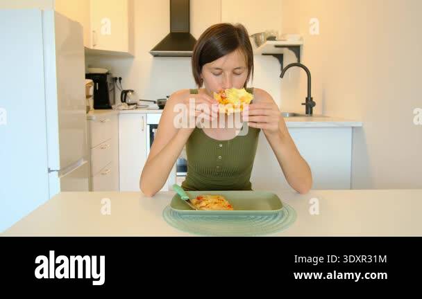 Woman eat pizza slice seated at white kitchen table , capturing simple ...