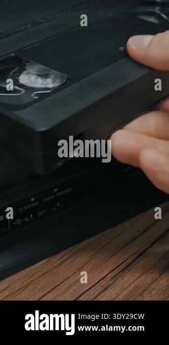 A close-up vertical shot shows a hand inserting a VHS tape into a VCR ...