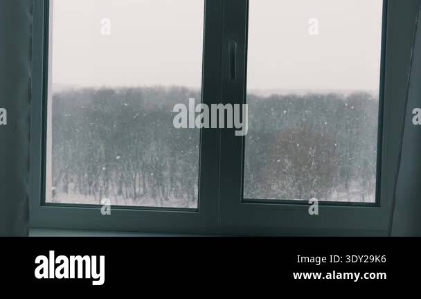 Heavy snowfall falls outside a window overlooking a winter forest ...