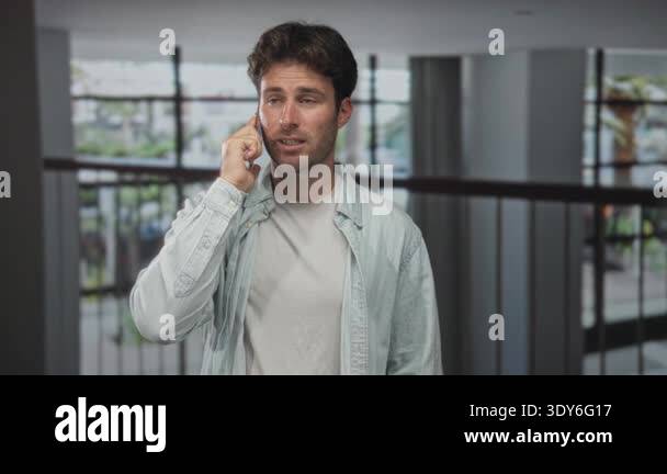 Man holding smartphone to ear in hotel lobby by glass railing, wearing ...