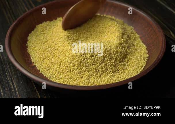 Yellow millet grains are seen in a wooden bowl as a wooden spoon stirs ...