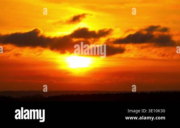 A bright sun descends through layers of dark clouds above a tree line ...