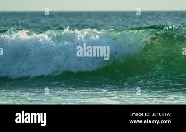 An ocean wave breaks gently at sunset, backlit by the sun. The scene ...