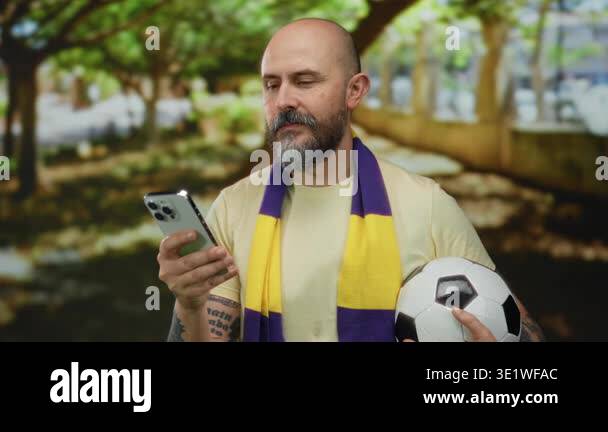 Bald man with beard holds smartphone and soccer ball, wearing yellow ...