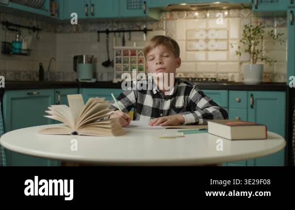 School boy studying at home, writing in a notebook while checking an ...