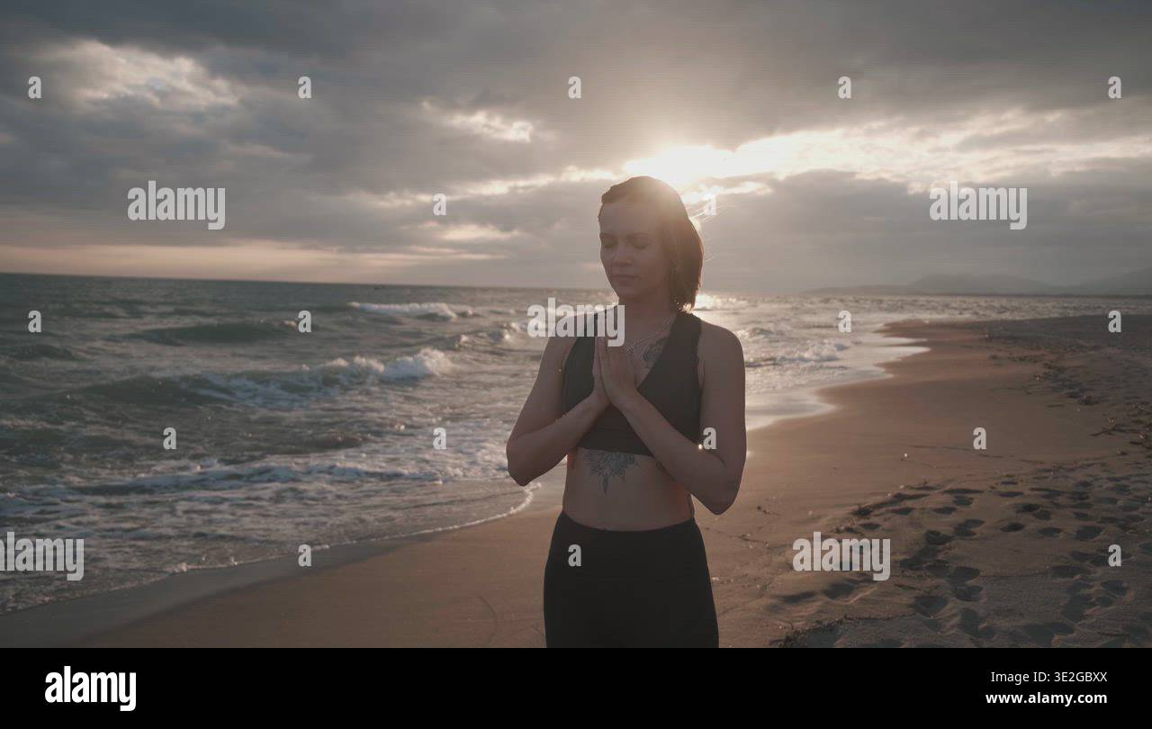 Woman is standing on the beach with close eyes and pose namaste. Sunset ...