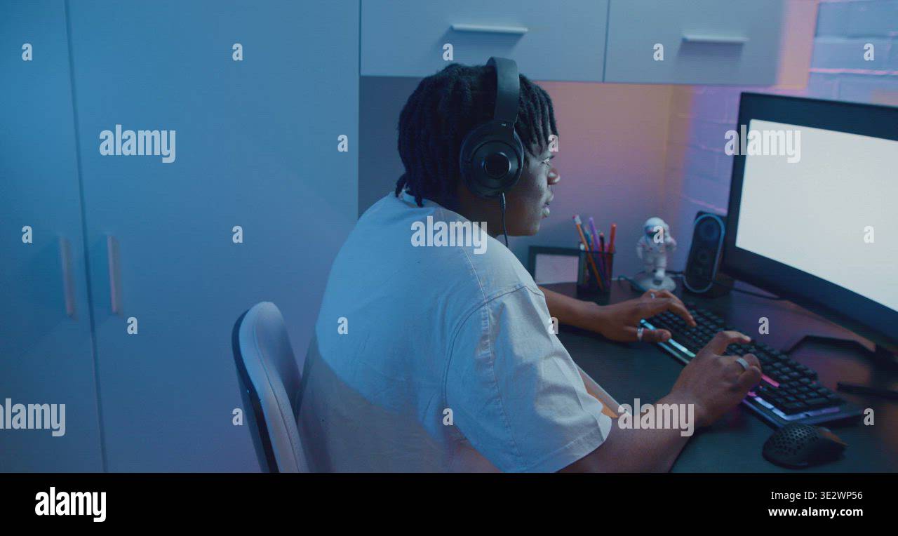Gamer focusing on computer screen wearing headphones in neon-lit room ...