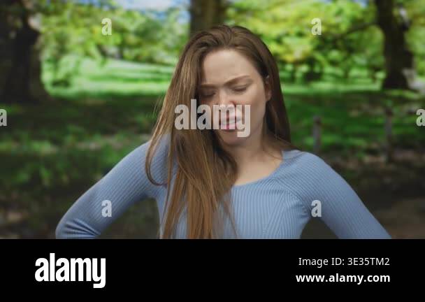 Woman frowns with a scrunched face and hands on hips in a sunlit park ...