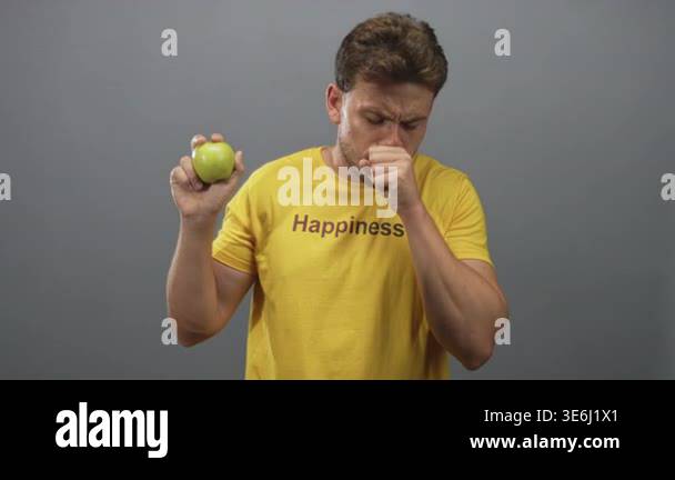 Man in yellow shirt holding green apple and coughing into fist while ...