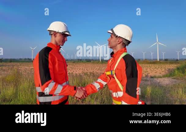 Two wind energy workers in orange safety gear shake hands at an onshore ...