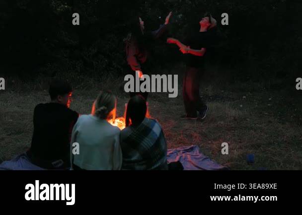At nightfall in the woods, teens gather around a fire. A girl and boy ...