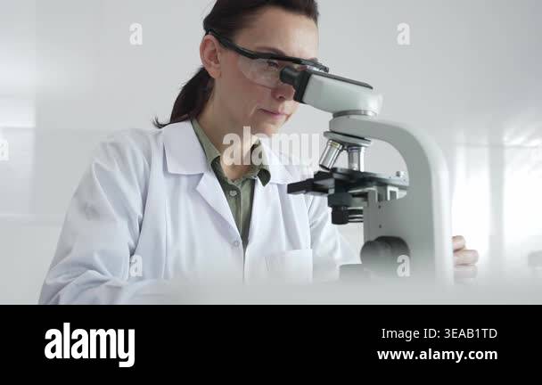 Female scientist wearing protective gear focusing on microscope while ...