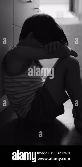 Young boy curled up indoors in black and white conveying vulnerability ...