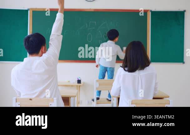 Young students raising hands while a boy writes on a chalkboard in a ...