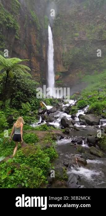 Woman walking toward Kapas Biru waterfall surrounded by lush tropical ...