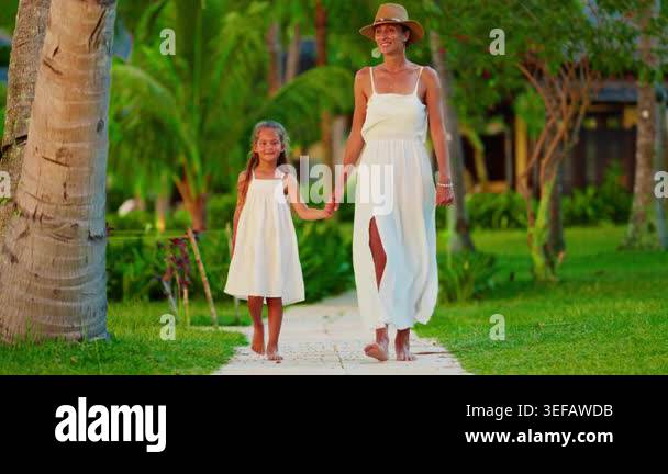 A mother and daughter on vacation, wearing white dresses, stroll hand ...
