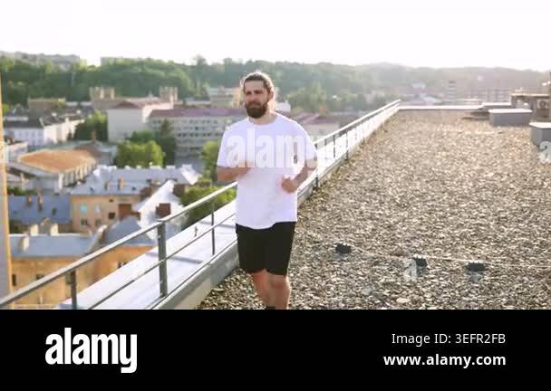 A bearded man with long hair runs on a gravel rooftop overlooking a ...