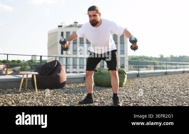 A bearded man with his hair tied back is performing a lateral raise ...
