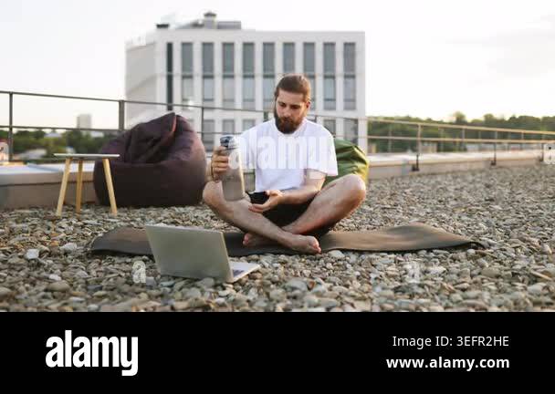 A bearded man with his hair in a bun sits cross-legged on a yoga mat on ...