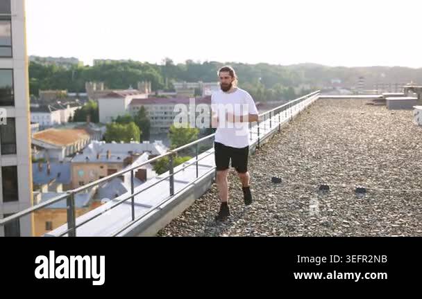 A man with a beard and bun hairstyle is jogging on a gravel rooftop ...