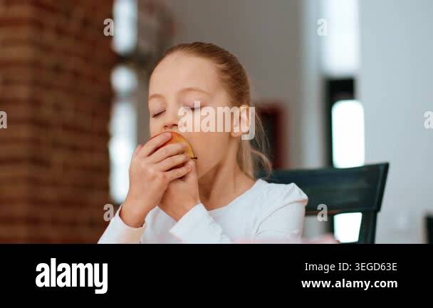 A girl with light skin and blonde hair bites into a red apple. She sits ...
