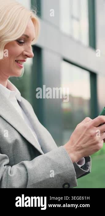 Portrait of happy blond business woman typing by mobile phone outdoors ...
