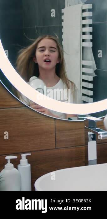 Pretty small girl dries her hair with hair dryer, singing and smiling ...