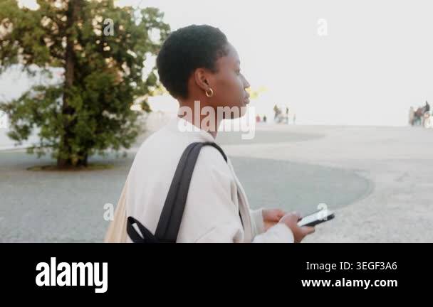 Young Black woman walking while using smartphone, focused on screen as ...