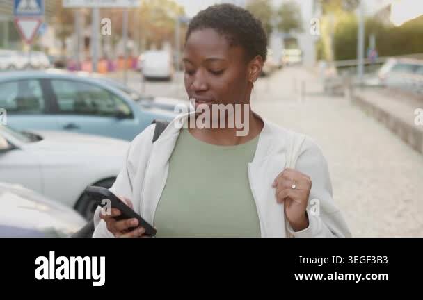 Smiling young black woman using smartphone while walking on sidewalk ...