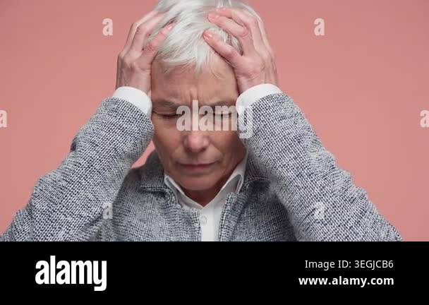 Older woman with short gray hair holds her head with closed eyes ...