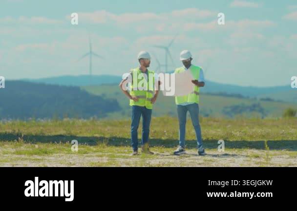 two engineers holding blueprint near turbines, sunny hilltop site ...