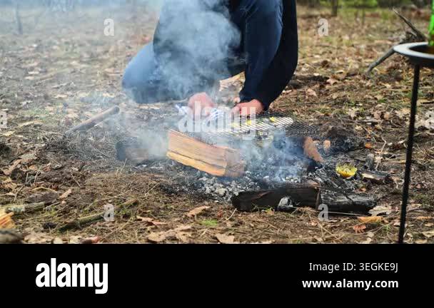 Close up of a hand holding a grill grate with fish over hot campfire ...