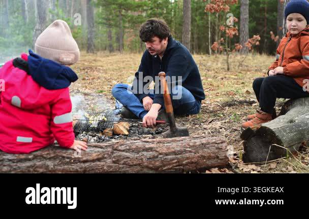 Wide shot of a man cooking fish on a grill over campfire coals while ...