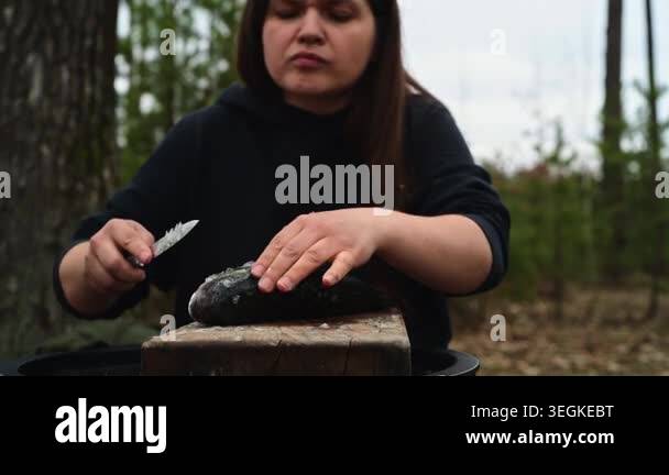 Woman sitting near a campfire in the forest cleaning a large sea bass ...