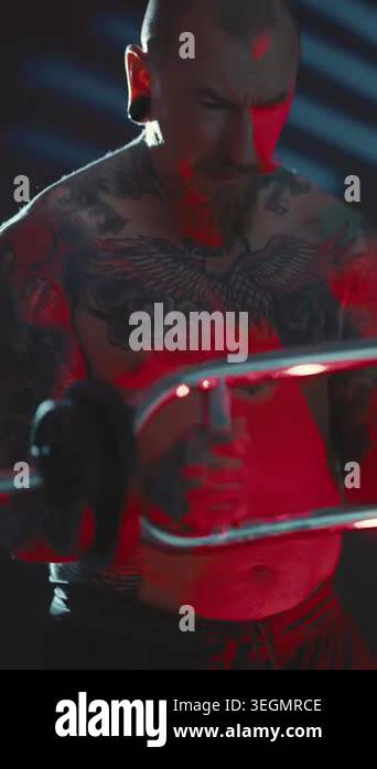 A man with tattoos is lifting weights in a gym. Bright lights create a ...