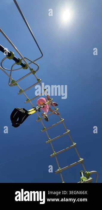 Woman climbing ladder against blue sky, pink outfit and harness visible ...