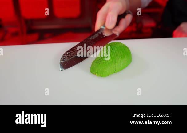 Chef hand slices fresh green avocado with a sharp knife on a white ...
