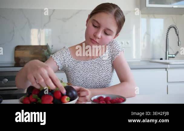 Girl enjoys selecting berries and grapes from bowls in a bright kitchen ...