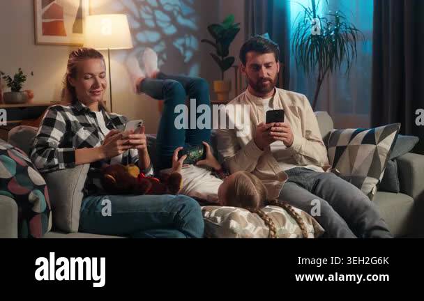 Family together on couch in evening living room using mobile phones ...