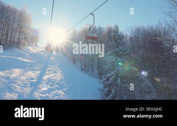 Back view of chair lift riding up to slope toward sunlight in winter ...