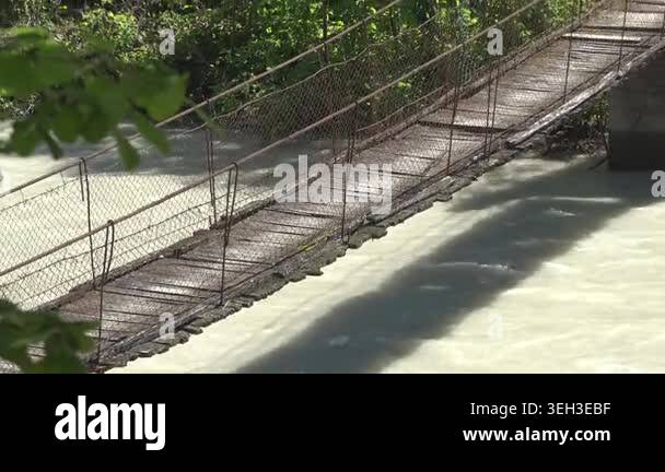 Wooden suspension bridge crossing wide flowing river within green rural ...