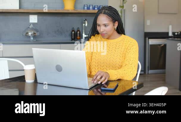 African American woman picking up coffee cup at kitchen table, typing ...
