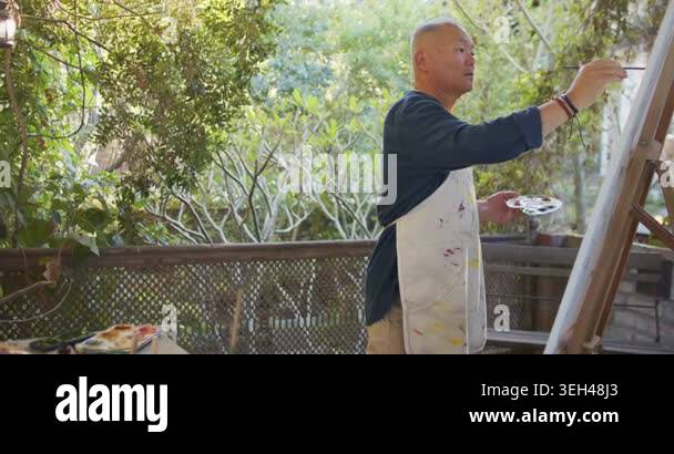 Senior Asian painter on porch painting at easel in stained apron ...