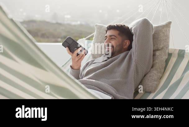 Indian man relaxing on terrace hammock, picking phone, smiling ...