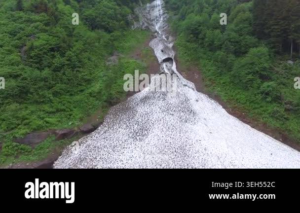 Aerial meltwater stream cuts through old avalanche snow on a forested ...