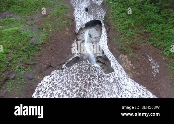 Aerial meltwater stream cuts through old avalanche snow on a forested ...