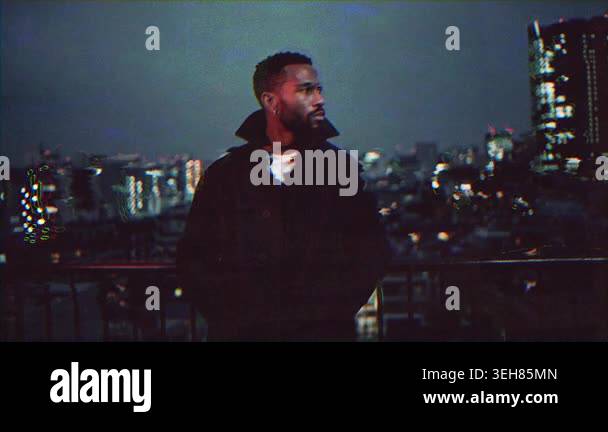 Man on rooftop at night wearing dark coat, city skyline blurred ...