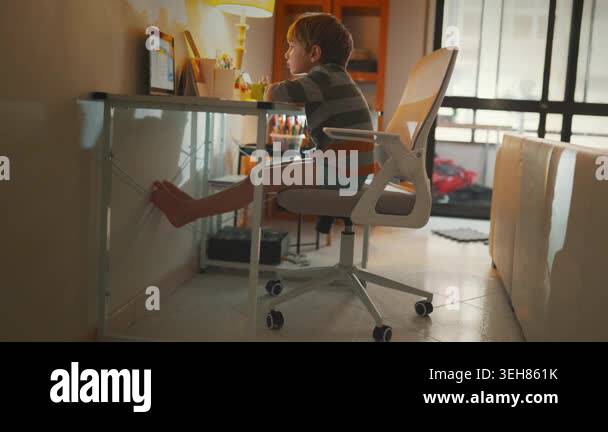 A young child sits at a desk in a home office. He focuses on the ...